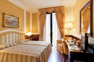 Quarto de hotel com uma cama grande, uma escrivaninha com cadeira, televisão e um quadro na parede.