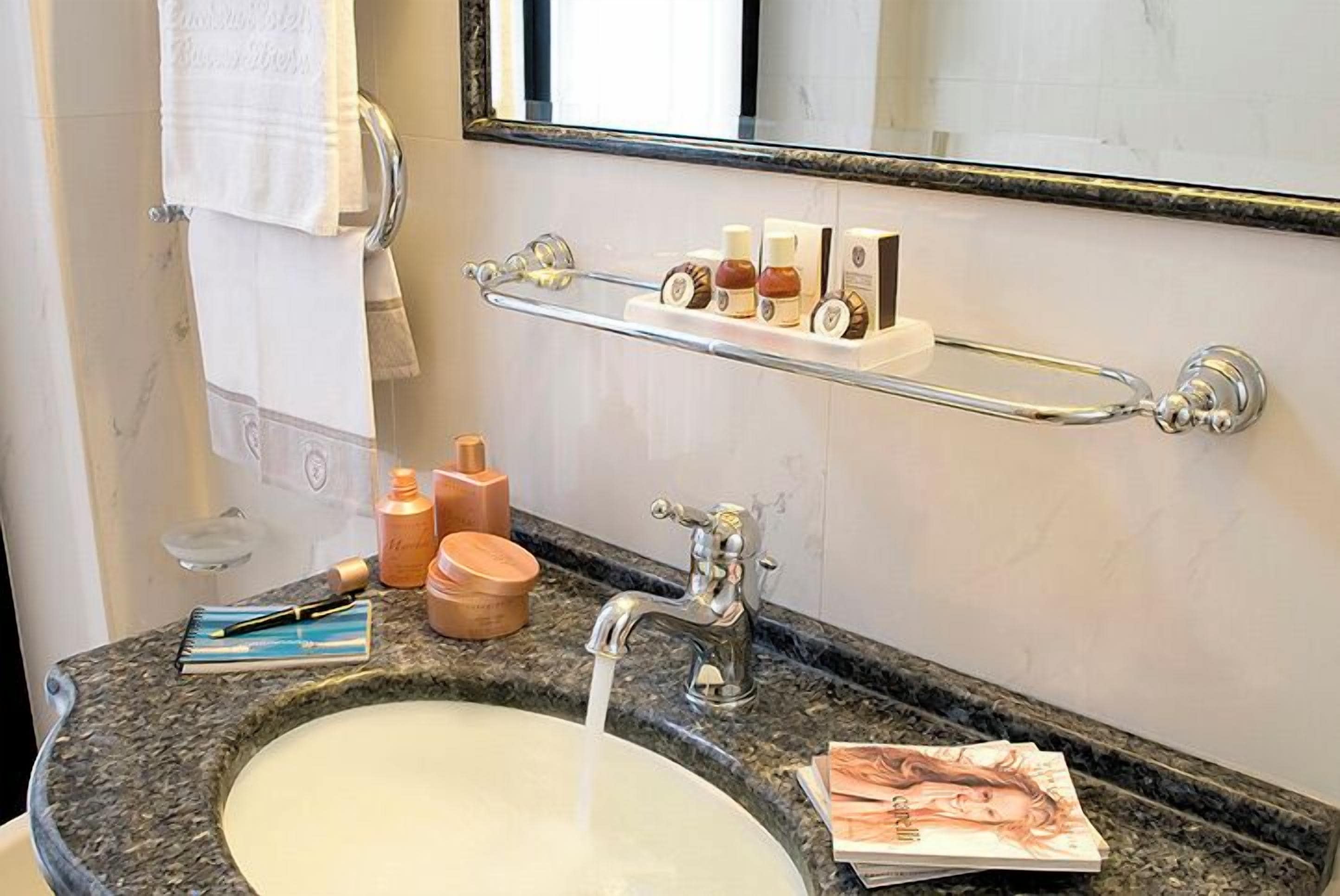 Casa de banho com lavatório de granito, um porta-toalhas com toalha branca, espelho e produtos de higiene numa prateleira.