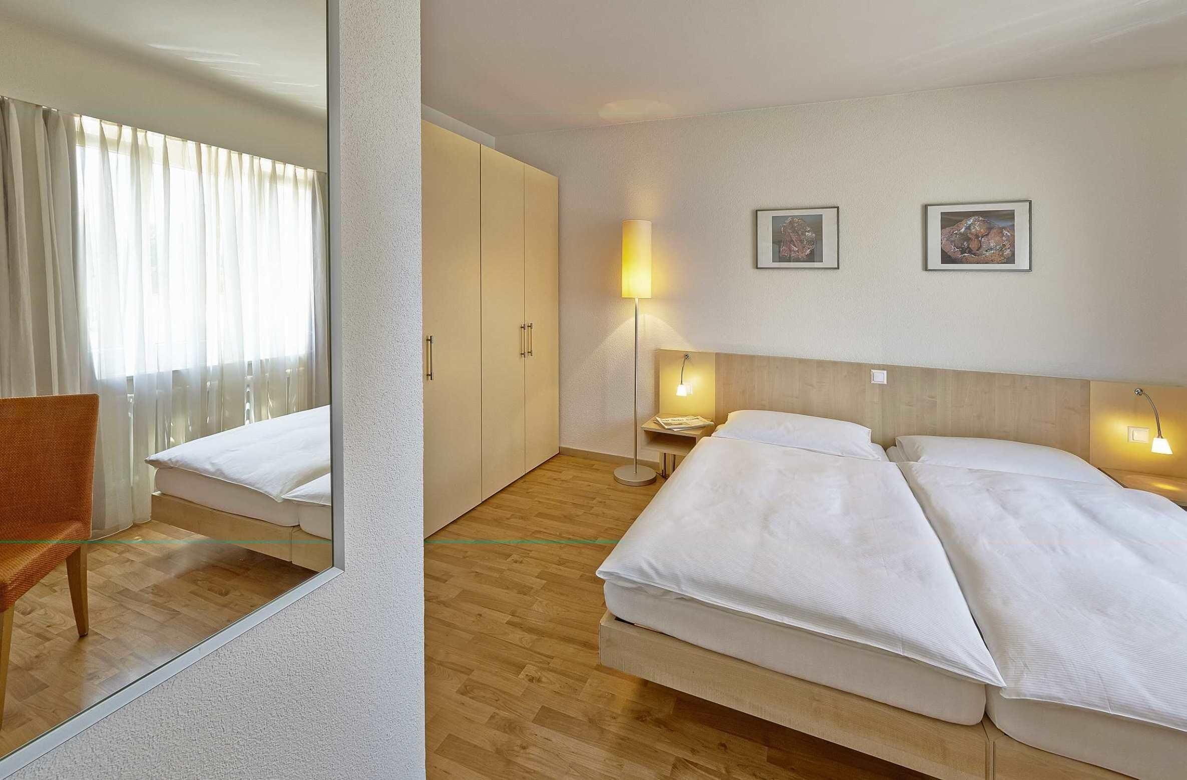 Quarto de hotel com cama, uma cadeira, mesa de cabeceira, abajur e duas fotos emolduradas na parede.
