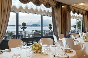 Restaurante com mesas postas para refeições, grandes janelas com vista para um lago e montanhas, e cortinas elegantes.