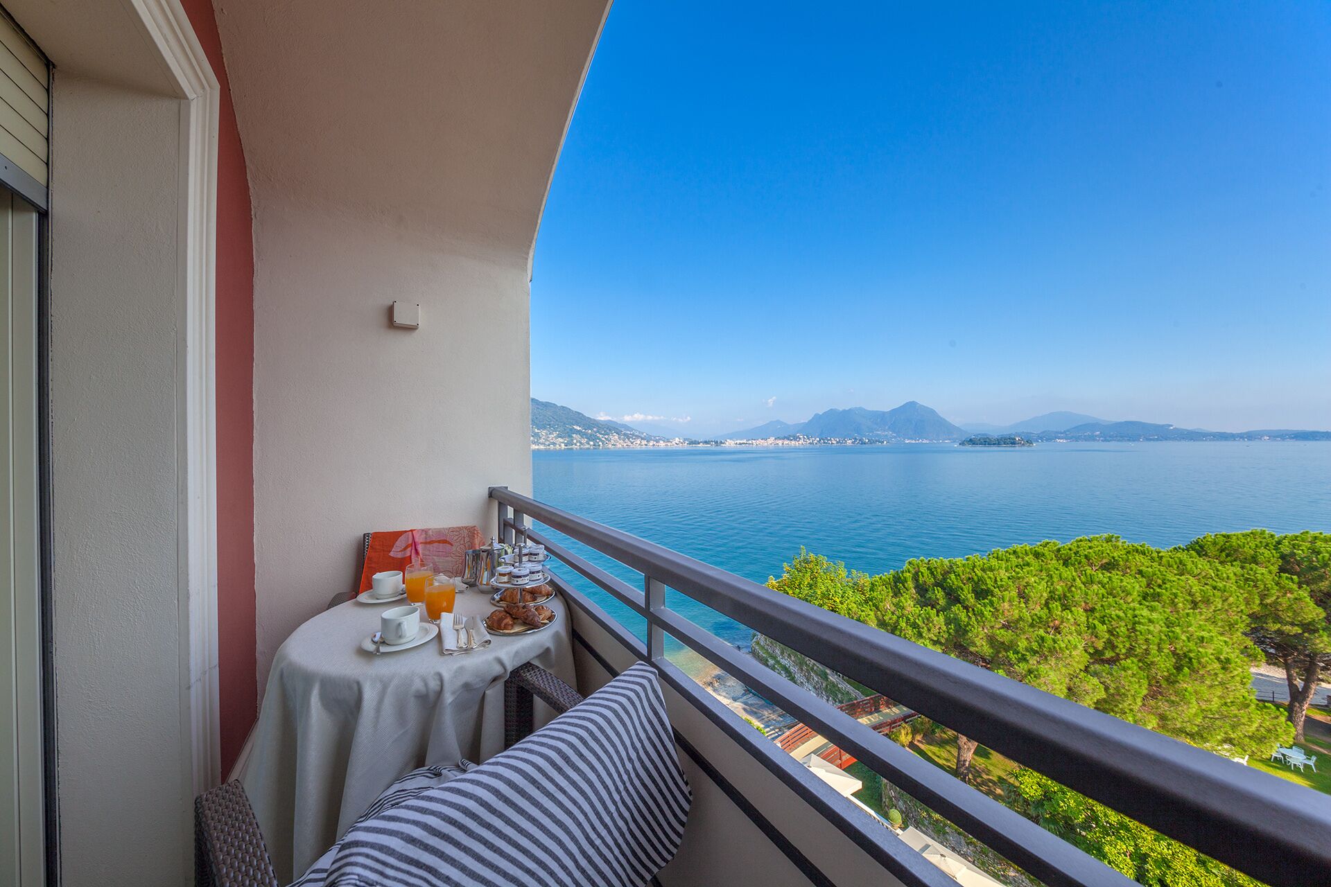 Varanda com mesa posta para café da manhã, com vista para um mar azul e límpido e montanhas ao longe.