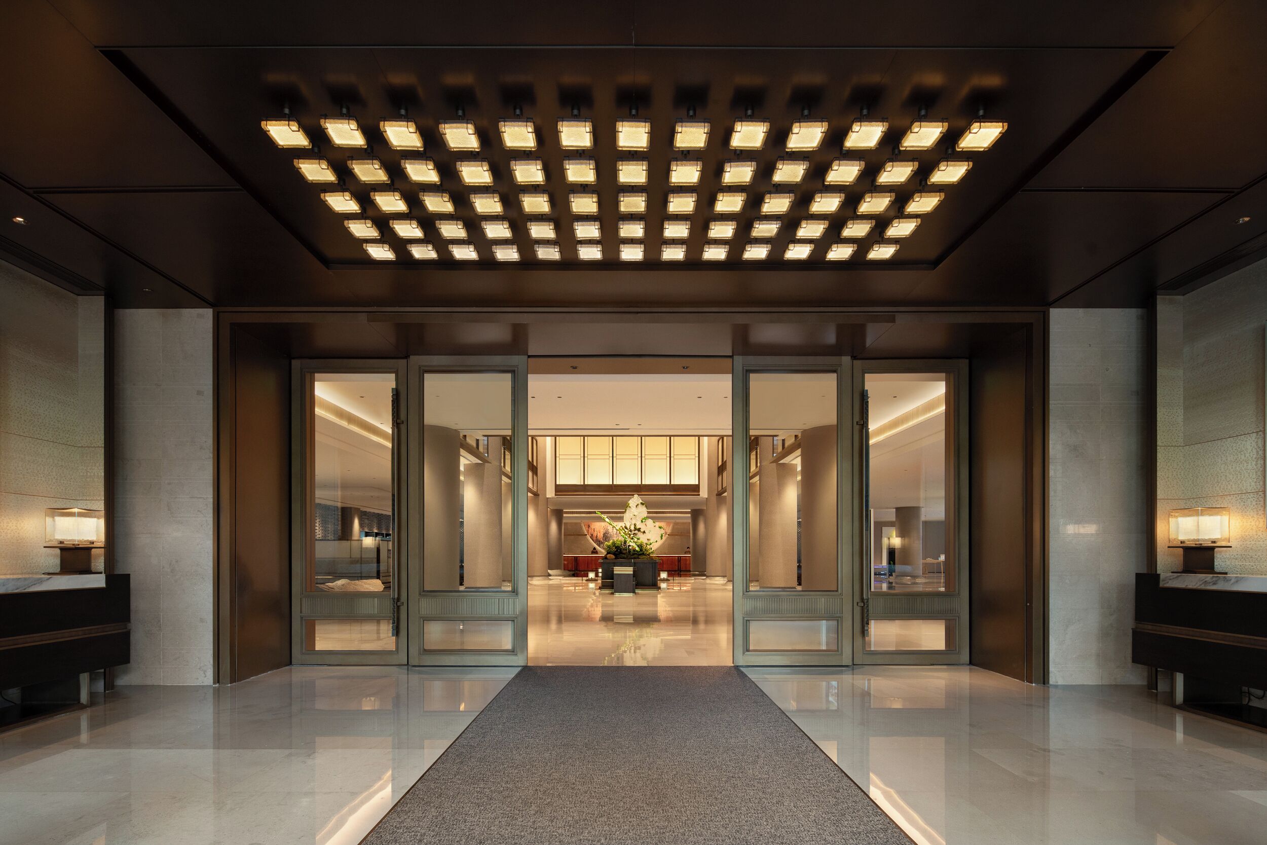 Um saguão de hotel com entrada por porta de vidro, um balcão de recepção central e piso coberto de carpete.