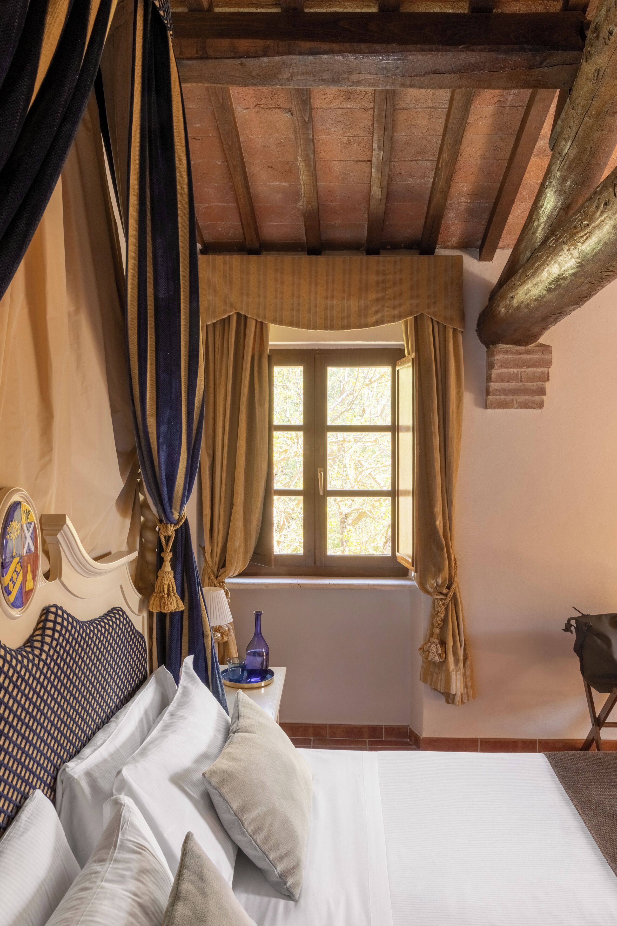 Um quarto com cama, janela com cortinas, teto de madeira e uma pequena mesa com abajur.