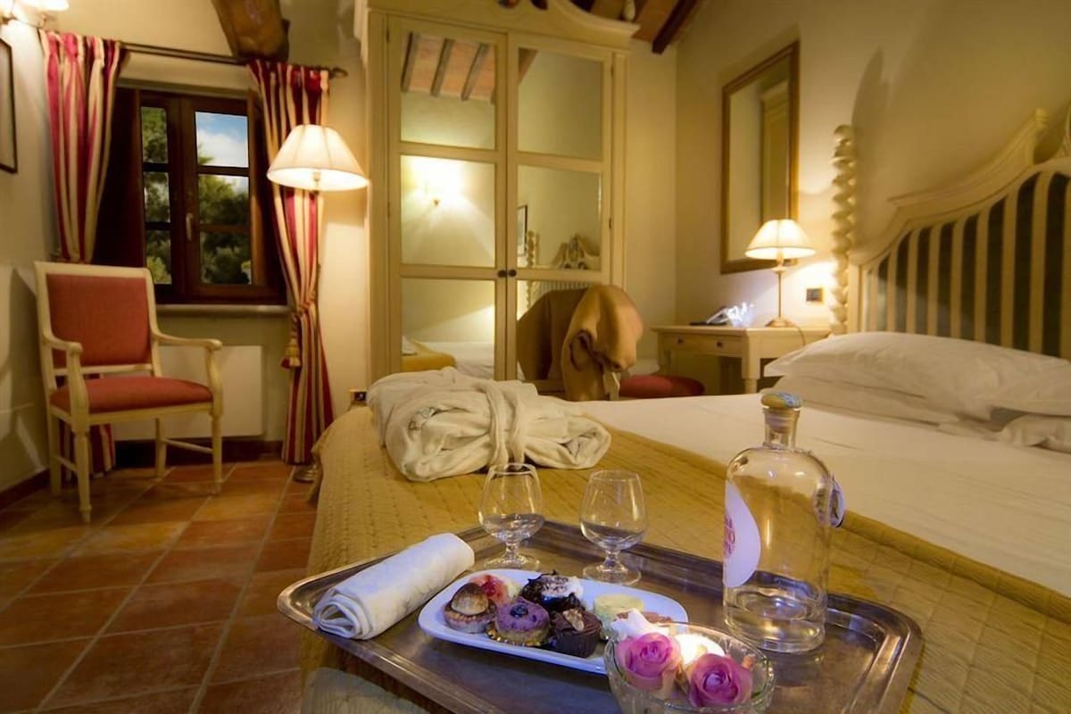 Quarto de hotel com cama, uma cadeira, mesa de cabeceira com luminária, bandeja com comida e bebida, e janela com cortinas.