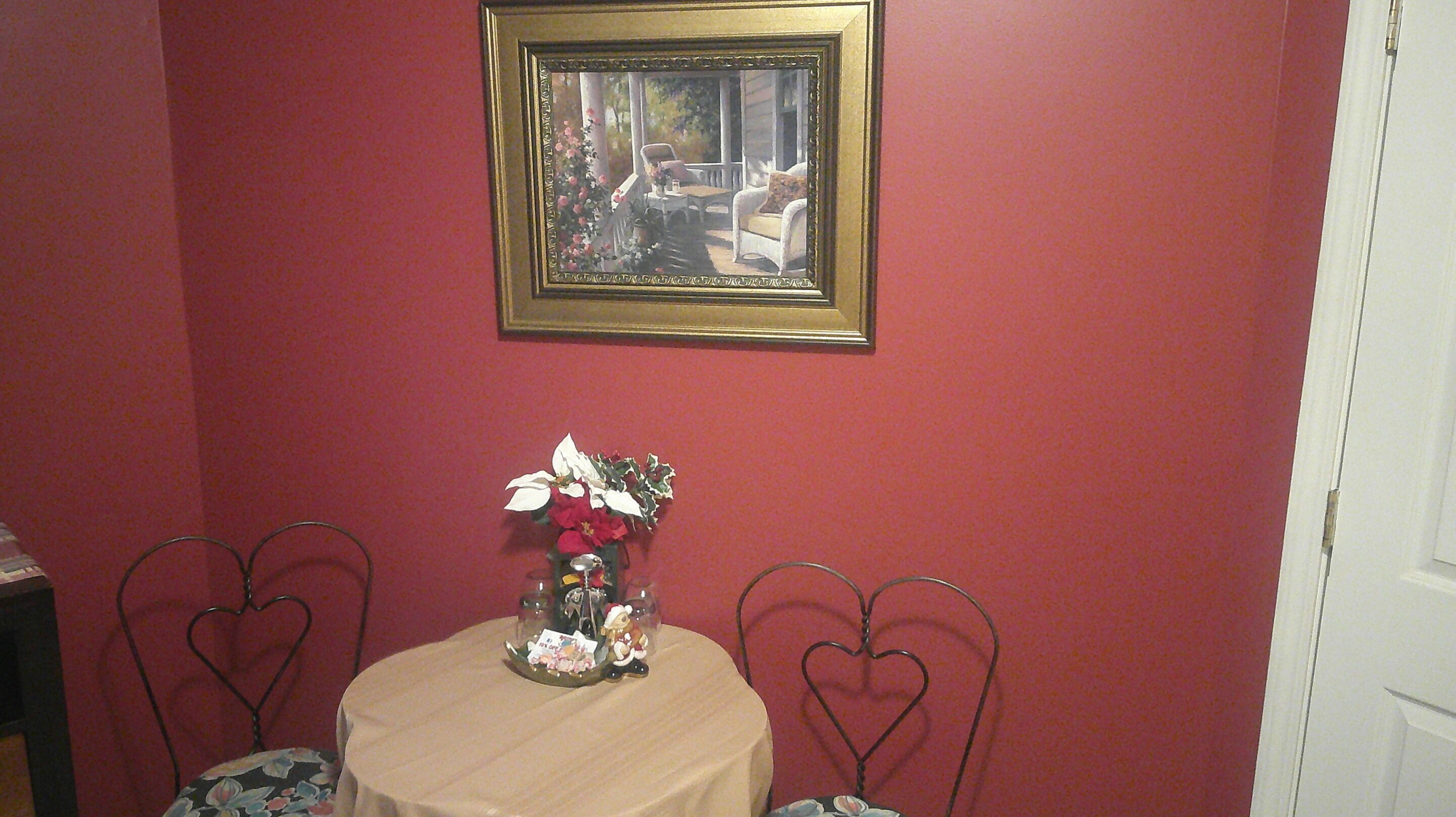 Uma área de jantar com uma mesa coberta por toalha bege, rodeada por cadeiras em formato de coração. Uma moldura com uma imagem está pendurada na parede acima da mesa.