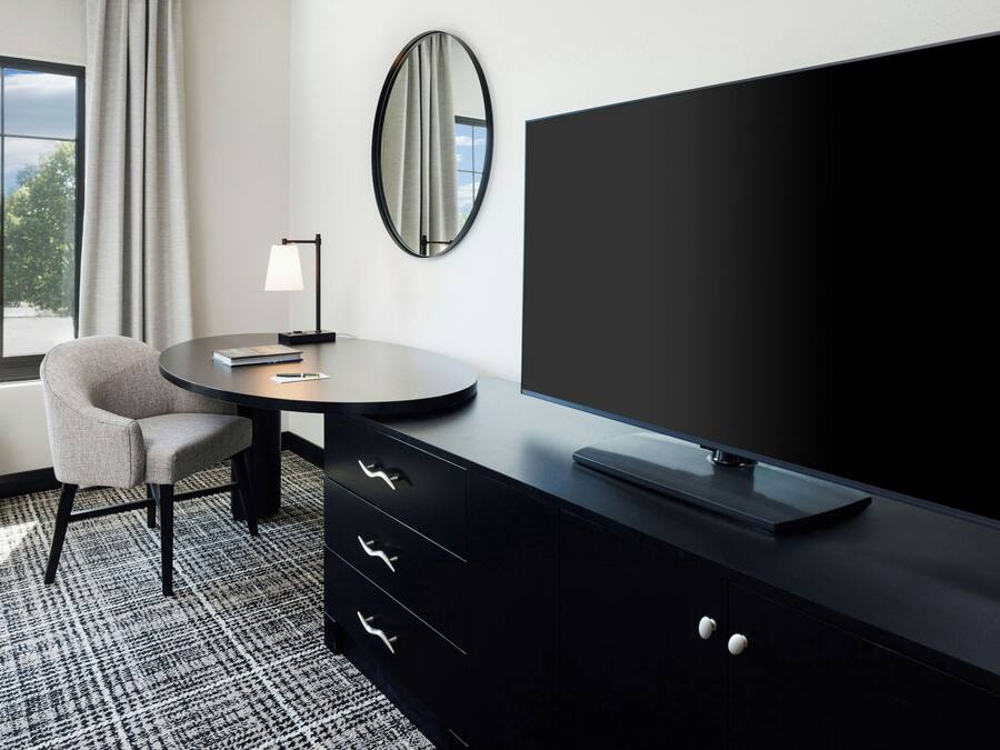 Quarto de hotel moderno com uma TV de tela plana grande, uma mesa redonda com luminária, uma cadeira e um espelho grande na parede.
