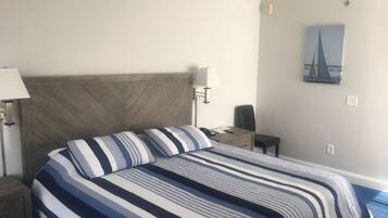 Quarto de hotel com uma cama grande, cabeceira de madeira, mesa de cabeceira com abajur, escrivaninha com cadeira e um quadro na parede.