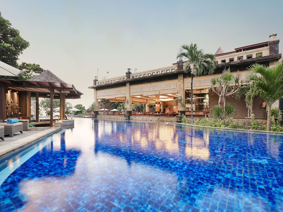 Uma piscina com superfície azulejada, área de estar coberta com espreguiçadeiras e um edifício com estilo arquitetônico tradicional ao fundo.