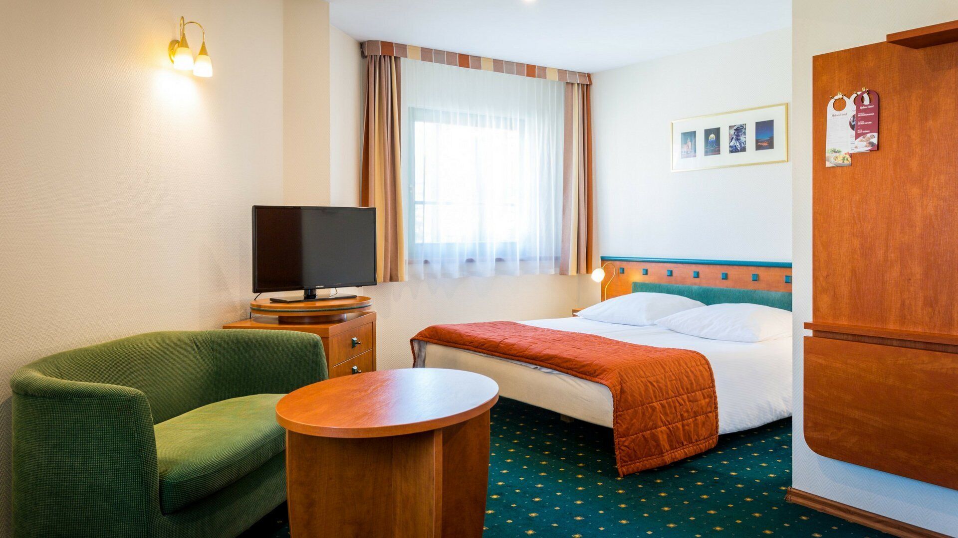 Quarto de hotel com cama, televisão, poltrona verde, mesa de madeira pequena e janela com cortinas.