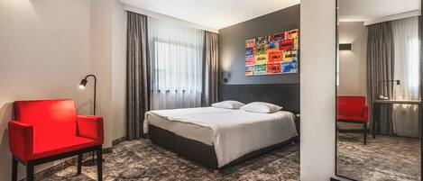 Quarto de hotel com cama, uma cadeira vermelha, um espelho grande e uma parede com arte colorida.