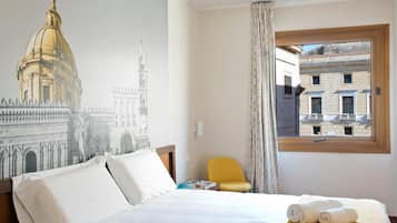 Quarto de hotel com cama, uma cadeira amarela, janela com vista para edifícios e um mural na parede de um edifício histórico.