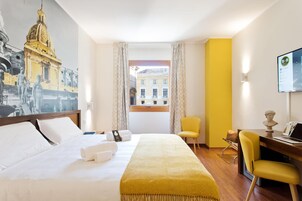 Quarto de hotel com cama, escrivaninha, cadeira, televisão e janela com vista para um edifício.