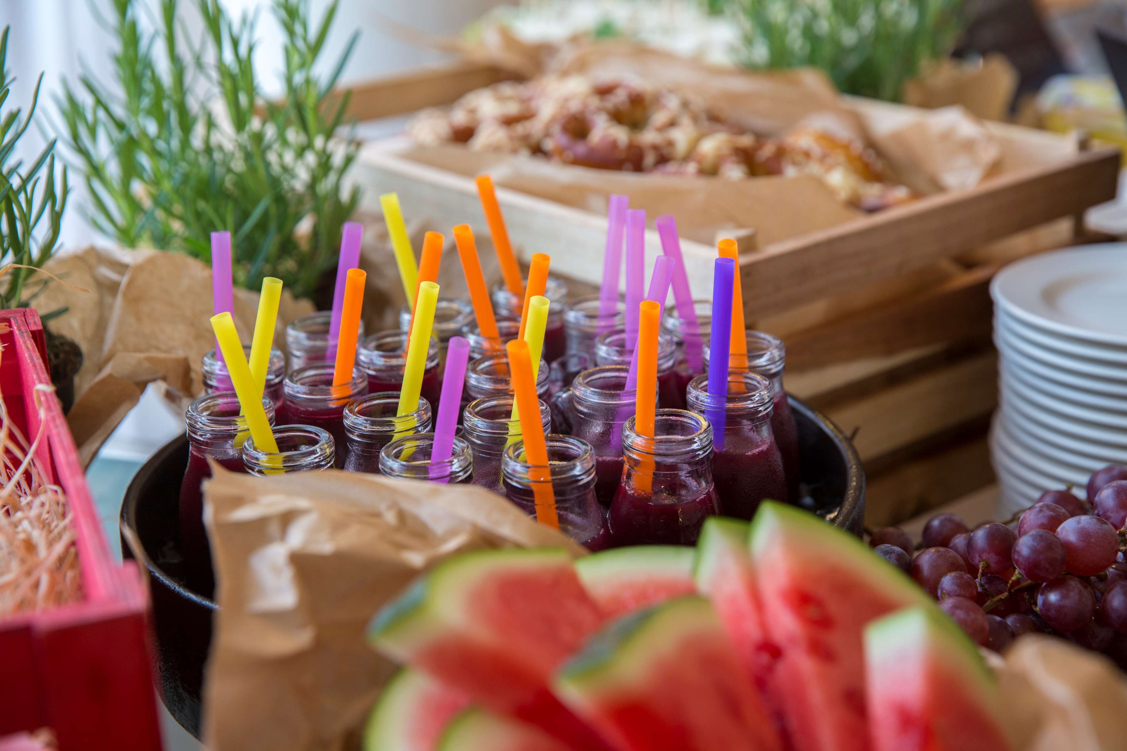 Uma mesa com diversos alimentos, incluindo potes de bebidas com canudos coloridos, uma fatia de melancia e uma tigela de uvas.