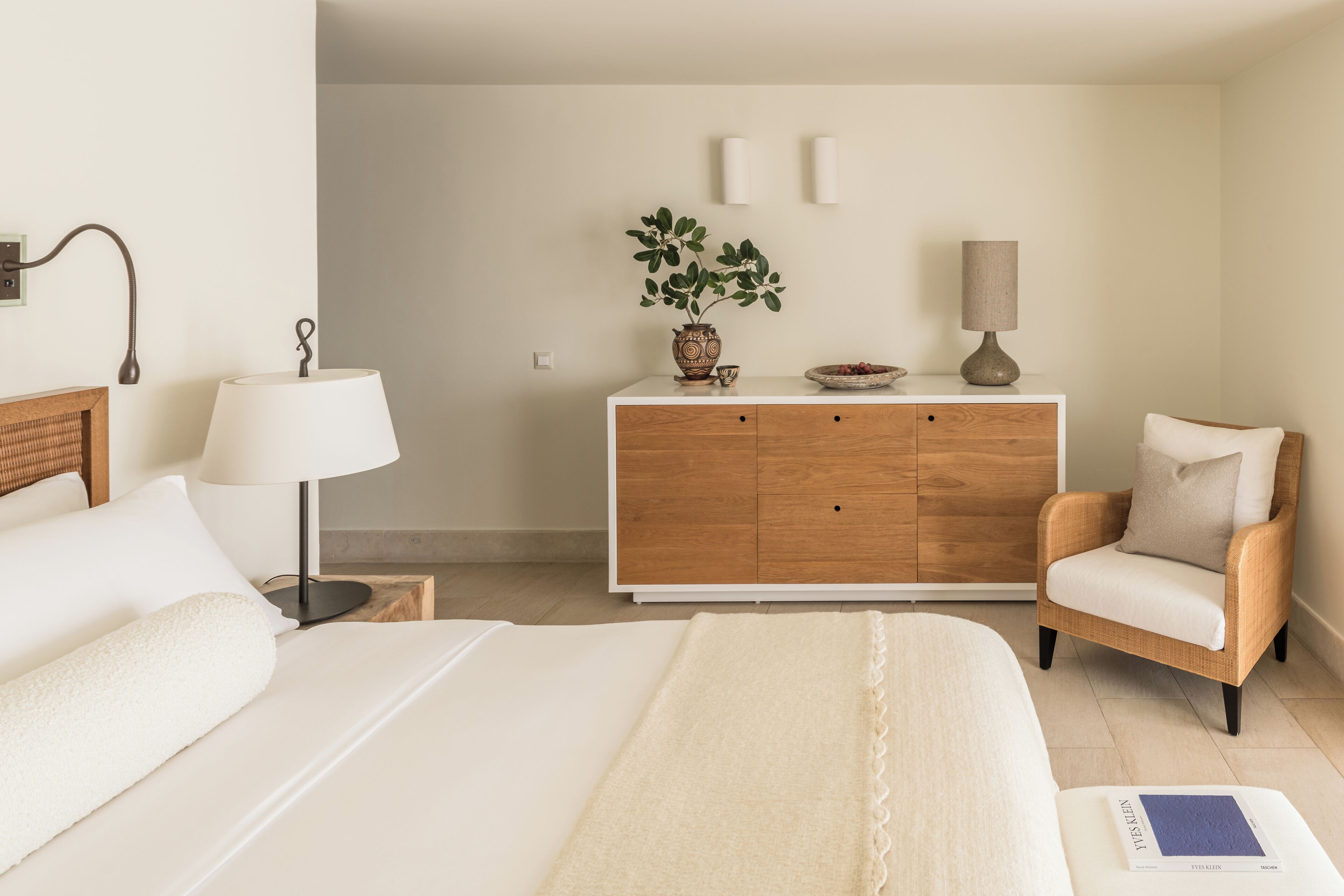 Quarto com cama, mesa de cabeceira, abajur, penteadeira, cadeira e uma pequena planta.