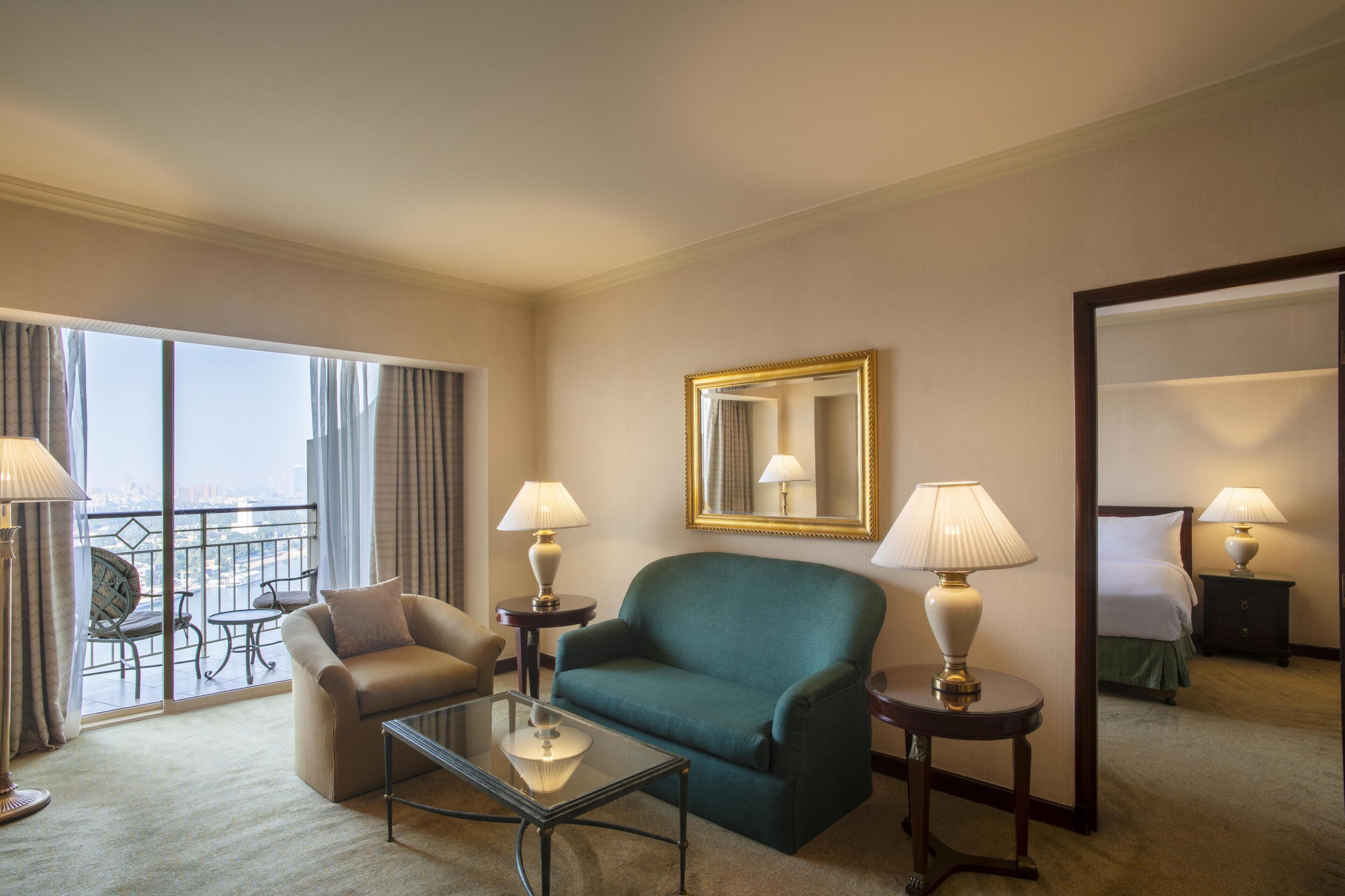 Quarto de hotel com varanda, uma poltrona verde, um sofá bege, uma mesa de centro de vidro e duas mesas de cabeceira com abajures.