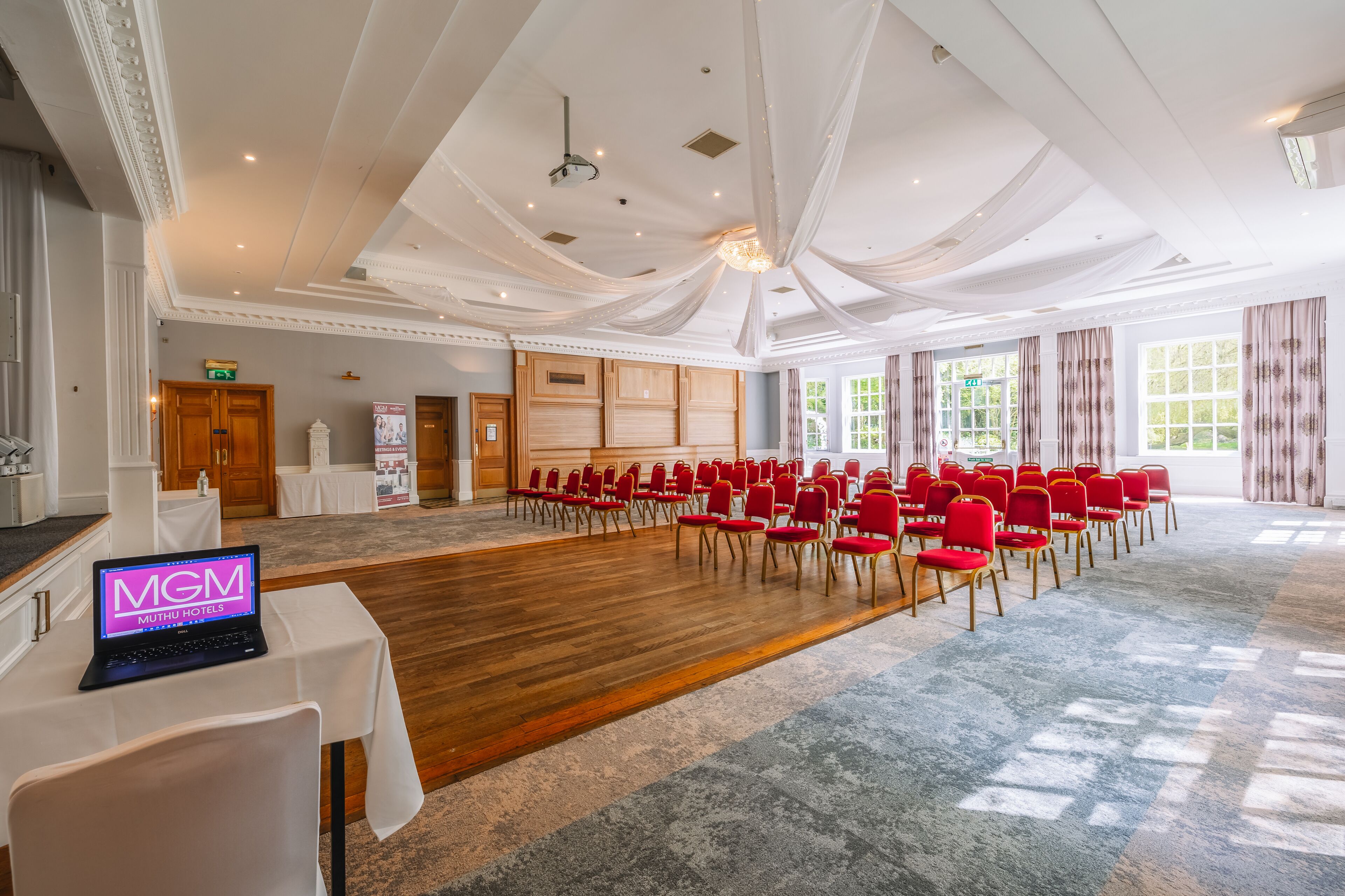 Uma sala de conferências ampla com fileiras de cadeiras vermelhas, piso de madeira e teto branco com tecido drapeado.