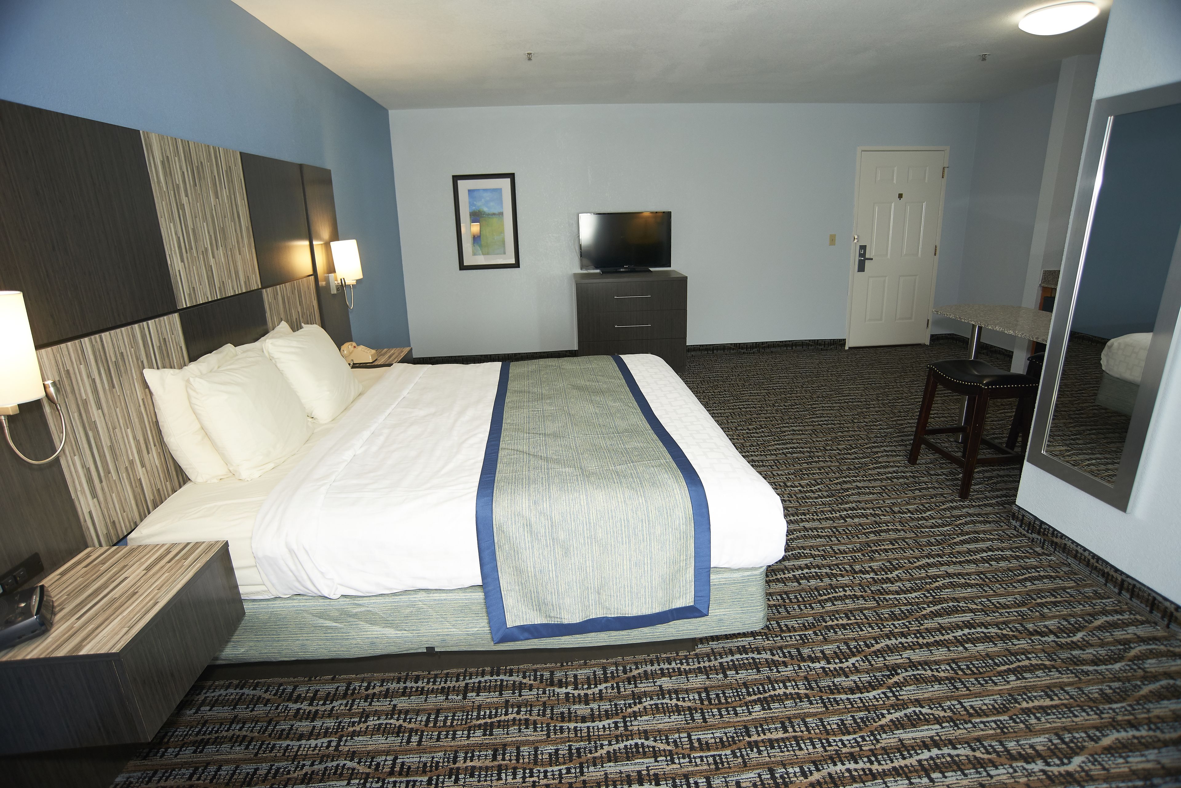 Quarto de hotel com uma cama grande, televisão de tela plana, escrivaninha com luminária, uma cadeira e uma porta.