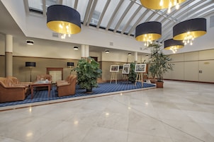 Um hall amplo com sofás de couro, mesa de centro, plantas em vasos, cavaletes com pinturas e luminárias pendentes modernas.