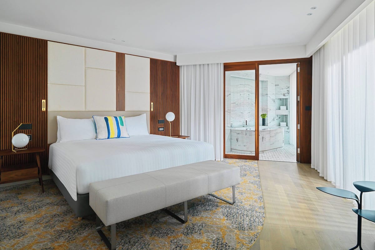 Quarto de hotel moderno com uma cama grande, um banco, uma escrivaninha e vista para o banheiro e varanda.