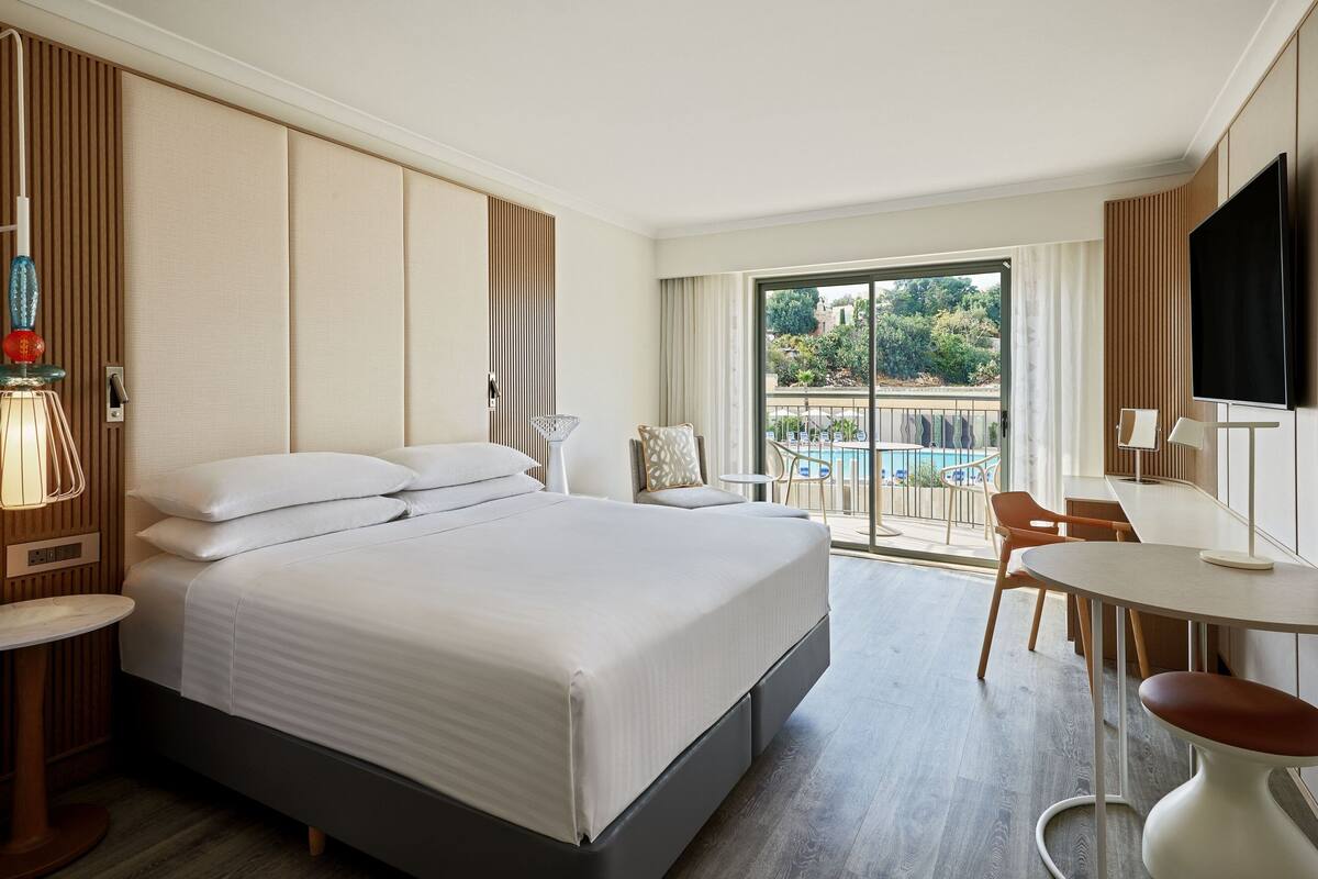 Quarto de hotel com cama grande, escrivaninha, cadeira, televisão e varanda com vista para a piscina.