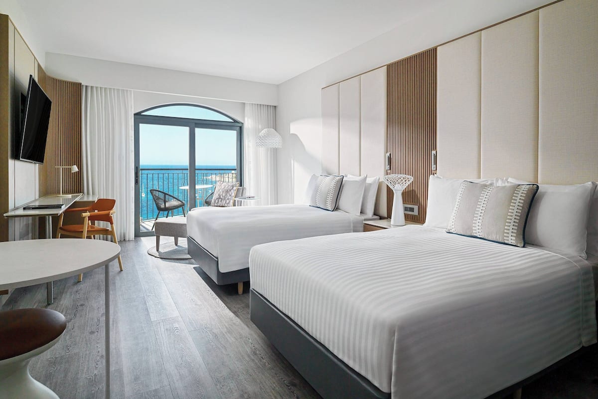 Quarto de hotel com duas camas, uma escrivaninha e uma cadeira. Há uma televisão na parede e uma varanda com vista para o mar.