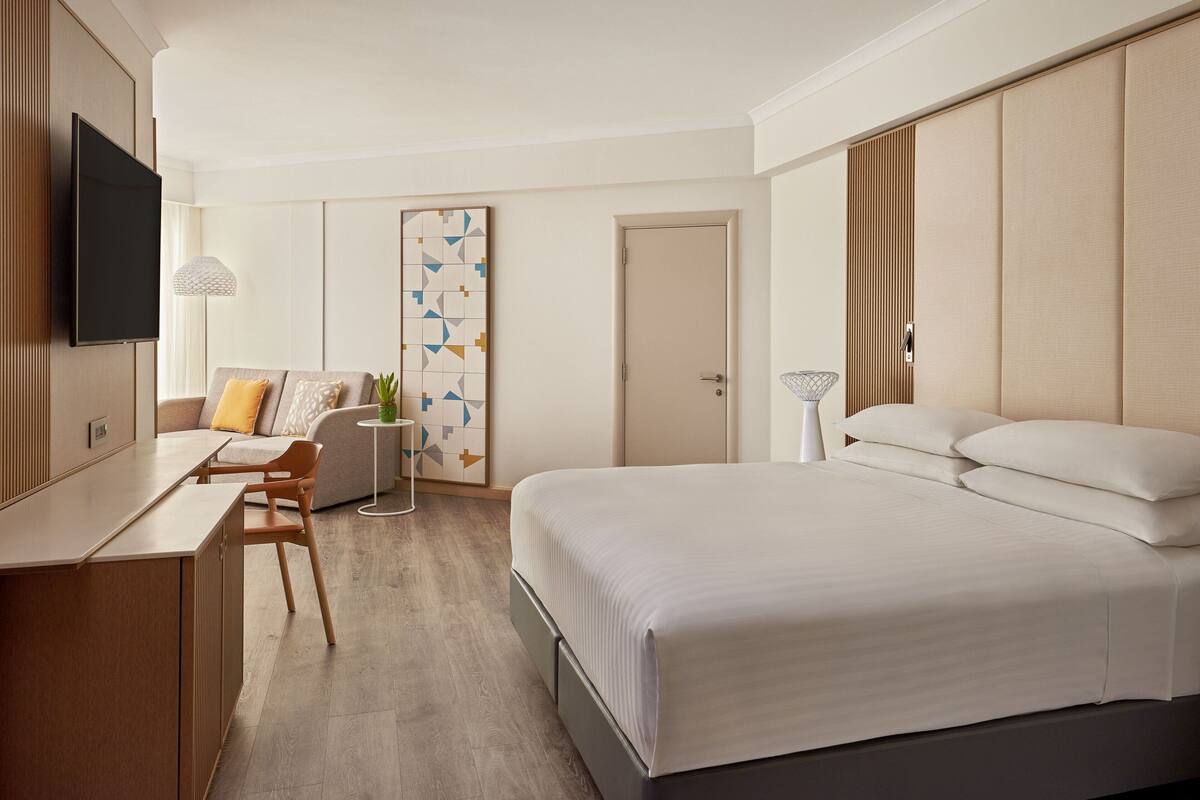 Quarto de hotel com cama, escrivaninha, cadeira, televisão e porta.