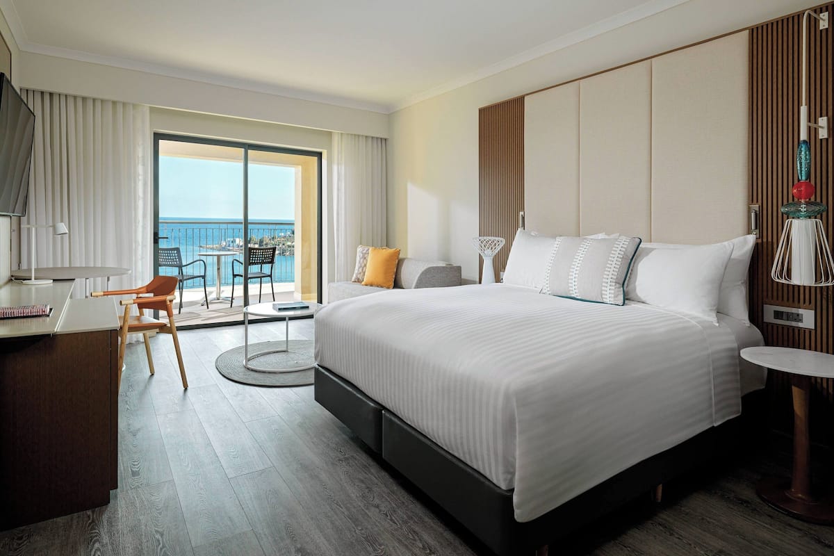 Quarto de hotel com uma cama grande, uma escrivaninha, uma cadeira e varanda com vista para o mar.