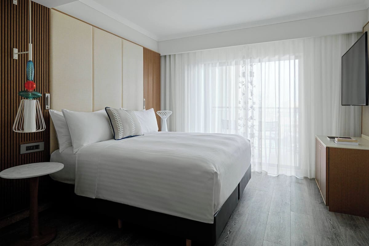 Quarto de hotel com uma cama grande, uma mesa de cabeceira, uma pequena mesa redonda, uma TV em um armário de madeira e uma janela com cortinas leves.