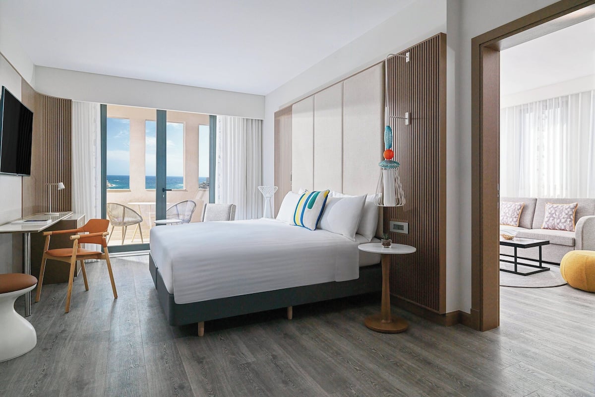 Quarto de hotel moderno com uma cama grande, uma mesa de trabalho e uma cadeira. Há uma varanda com vista para o oceano.
