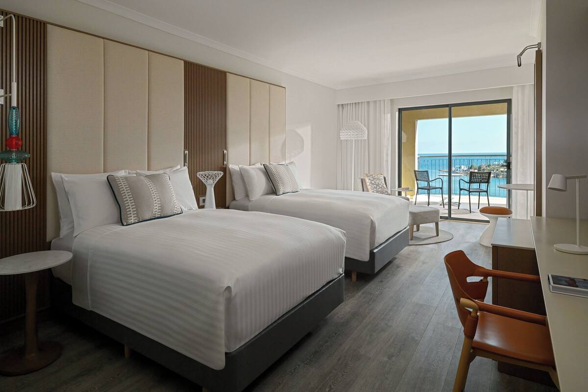 Quarto de hotel com duas camas, uma mesa de trabalho e varanda com vista para o mar.
