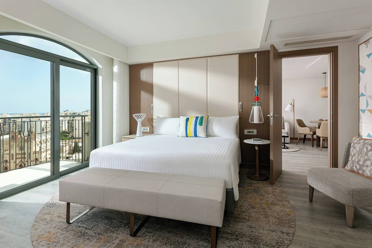 Quarto de hotel moderno com uma cama grande, um banco, vista para a cidade e um espaço de refeições.