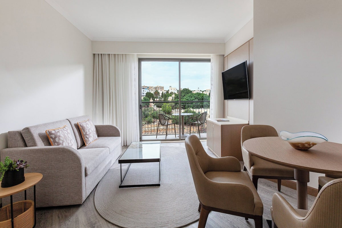 Uma sala de estar moderna com sofá, mesa de centro e área de jantar. Uma janela grande oferece vista da cidade.