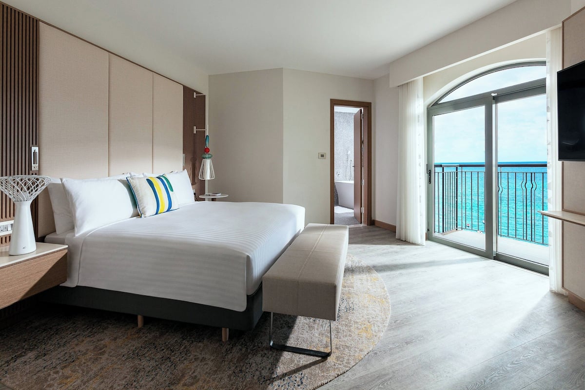 Quarto de hotel moderno com uma cama grande, um banco, uma televisão e uma varanda com vista para o mar.