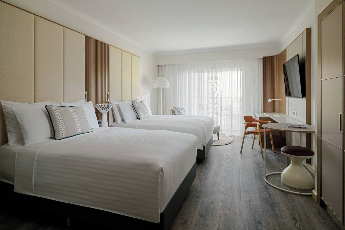 Quarto de hotel com duas camas, uma escrivaninha, uma cadeira, uma televisão e uma janela grande com cortinas.