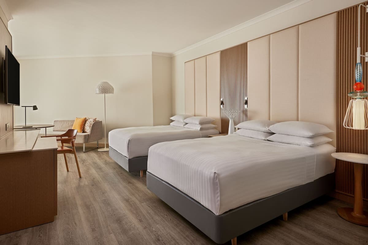 Quarto de hotel com duas camas, uma escrivaninha, uma cadeira e uma televisão presa à parede.