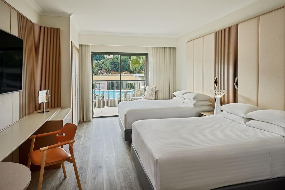 Quarto de hotel com duas camas, uma escrivaninha, uma cadeira, televisão e varanda com piscina.