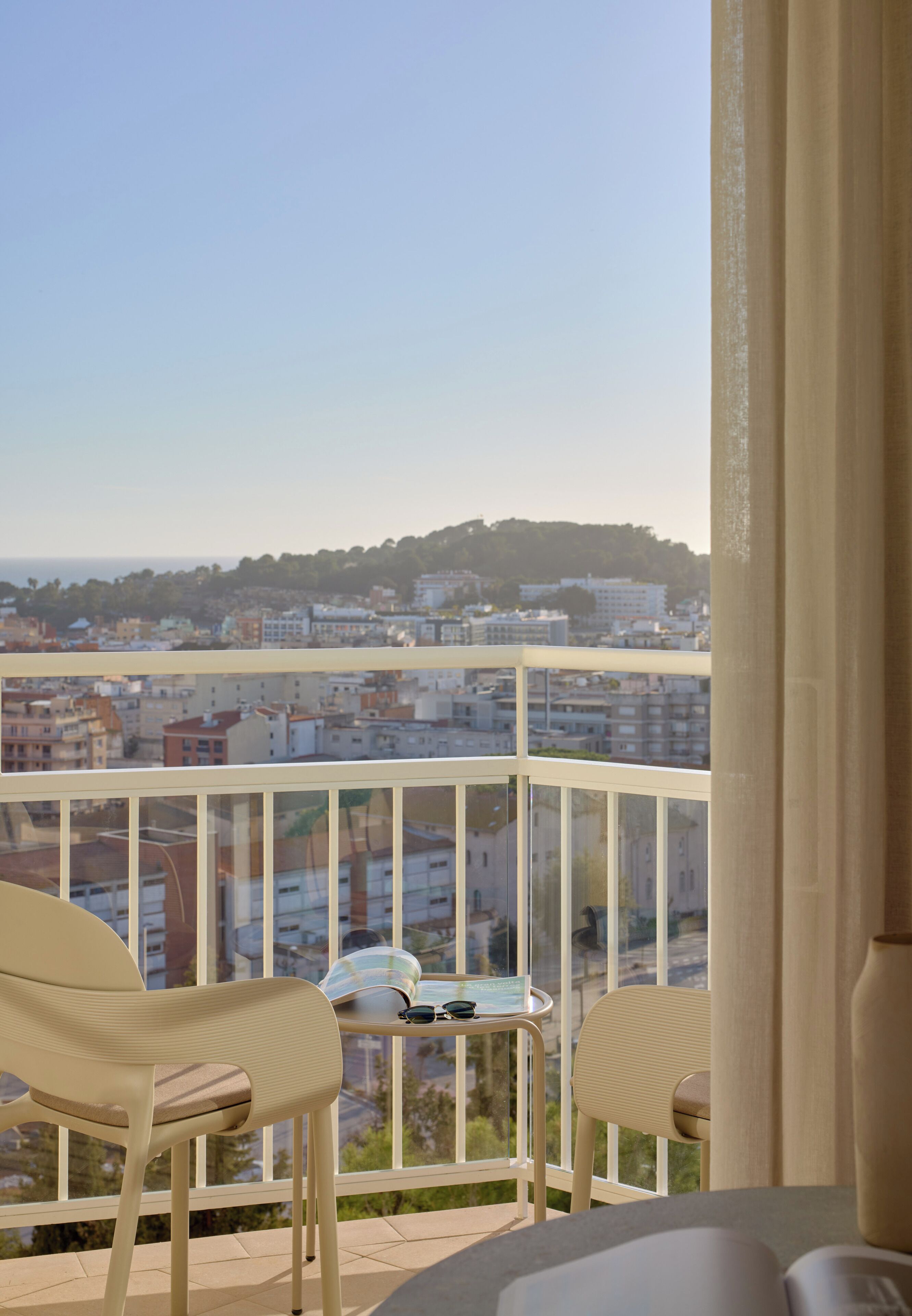 Varanda com duas cadeiras e uma mesa, com vista para um horizonte urbano com edifícios e árvores.