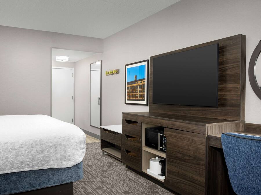 Quarto de hotel com cama, televisão, espelho e secretária pequena com gavetas.