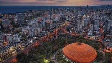 Paisagem urbana ao anoitecer, com uma estrutura em cúpula laranja destacada, rodeada por vegetação e vias.