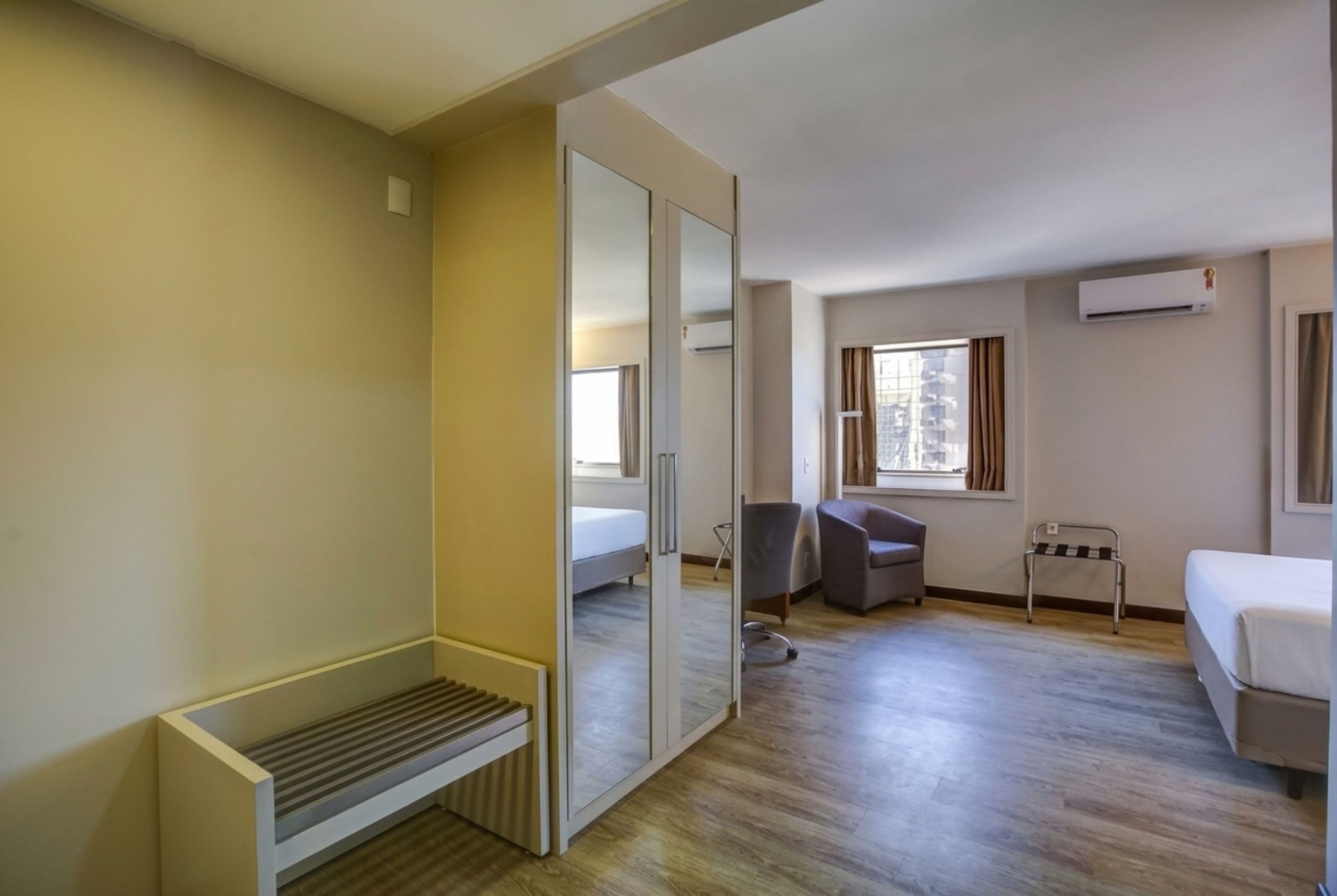 Quarto de hotel com cama, escrivaninha, cadeira, radiador e janela com cortinas.
