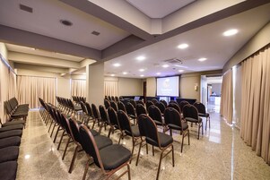 Uma sala de conferências com fileiras de cadeiras, uma tela de projetor e uma tela para apresentação.