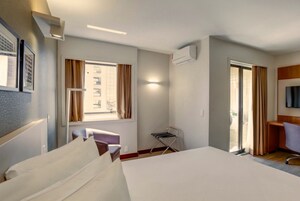 Quarto de hotel com cama, poltrona, televisão e janela com cortinas.
