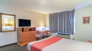 Quarto de hotel com cama, escrivaninha, cadeira, televisão, cômoda e janela com cortinas.