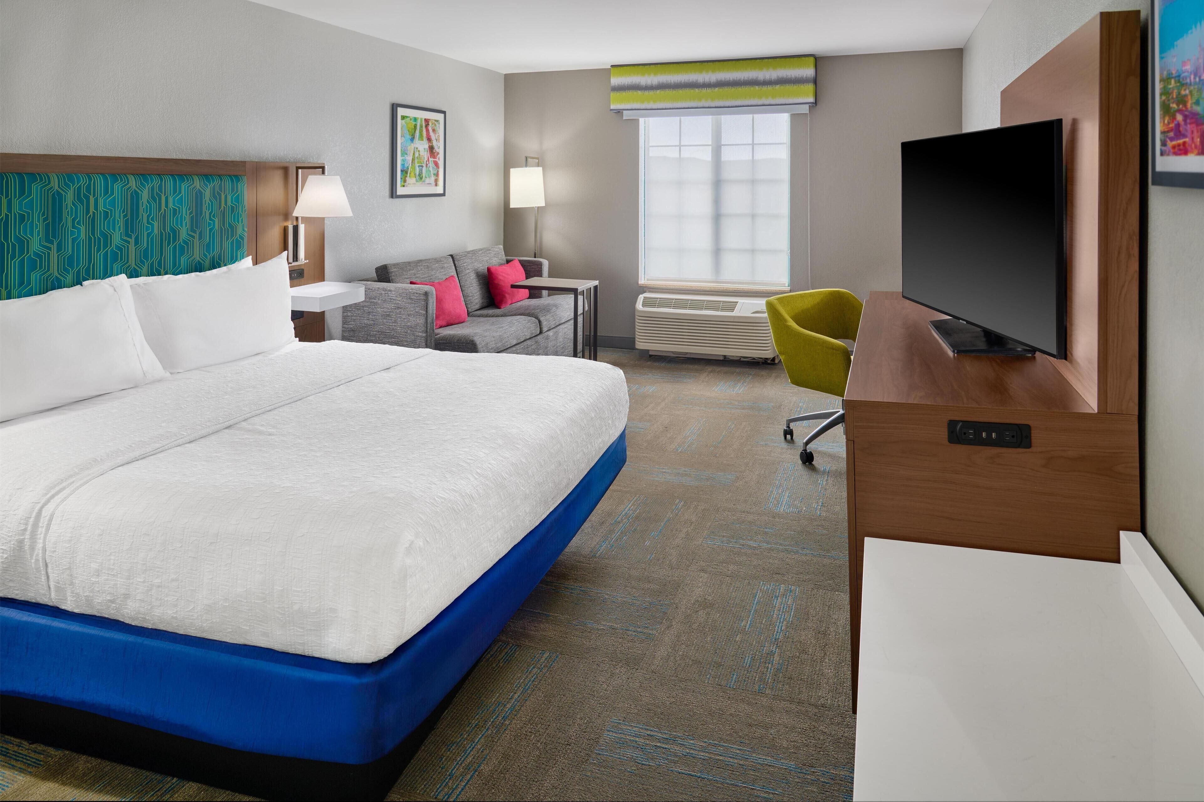 Quarto de hotel com uma cama grande, uma escrivaninha com televisão de tela plana, uma cadeira, uma mesinha e uma janela com persianas.