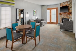 Quarto de hotel com área de refeições, sala de estar com lareira e parede de destaque em pedra.