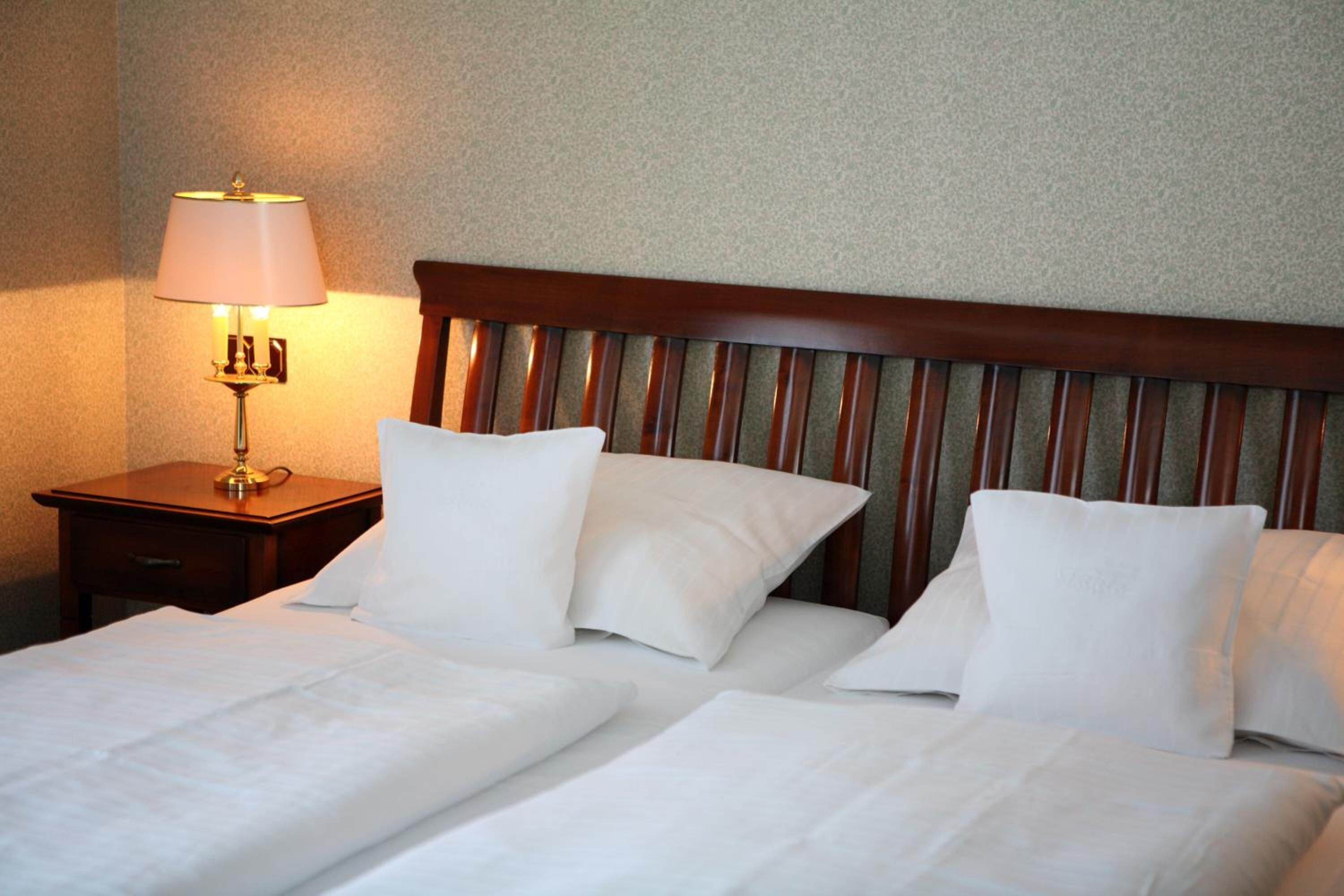 Quarto de hotel com cabeceira de madeira, duas camas com roupa de cama branca e mesa de cabeceira com abajur.