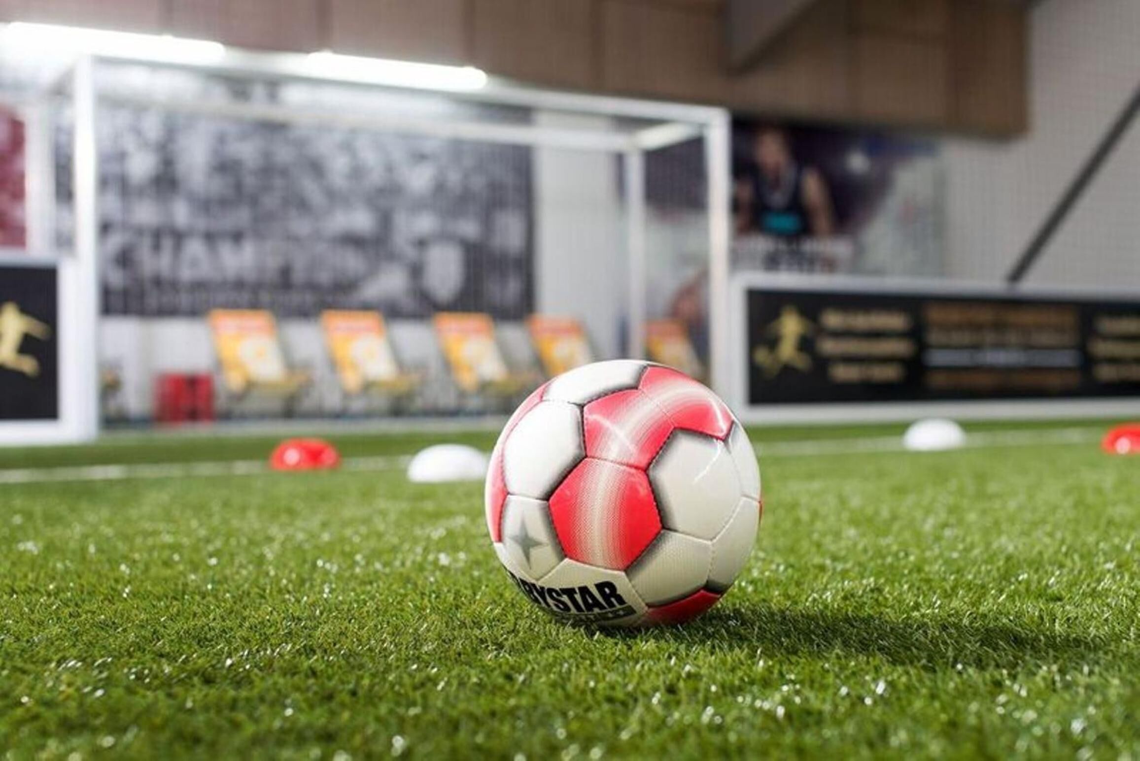 Uma bola de futebol em grama sintética, com um fundo desfocado de um gol e anúncios.