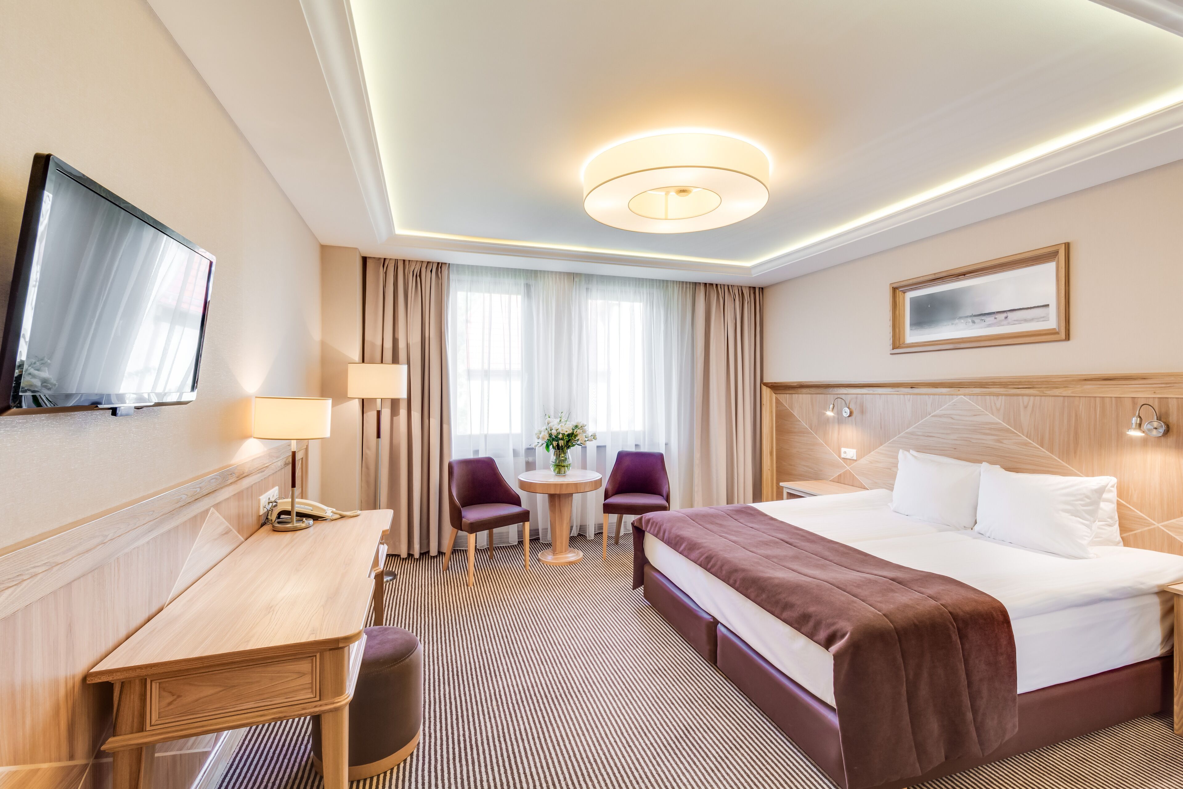 Quarto de hotel com uma cama grande, uma escrivaninha, duas cadeiras, uma televisão e uma janela com cortinas.