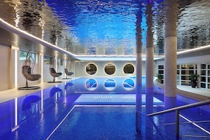 Piscina interna com vista clara para o teto, área de estar com cadeiras e área lateral à piscina com guarda-corpo.