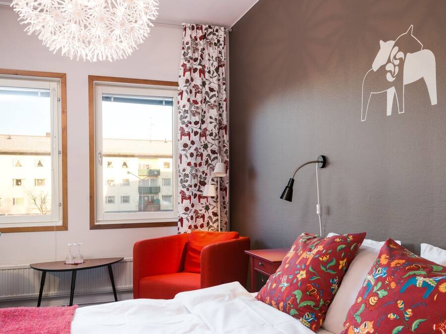 Quarto de hotel com uma cama grande, uma poltrona vermelha, uma mesinha e um abajur decorativo.